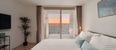 Quarto de hotel com uma cama grande, televisão, planta em vaso e vista para o mar ao pôr do sol.