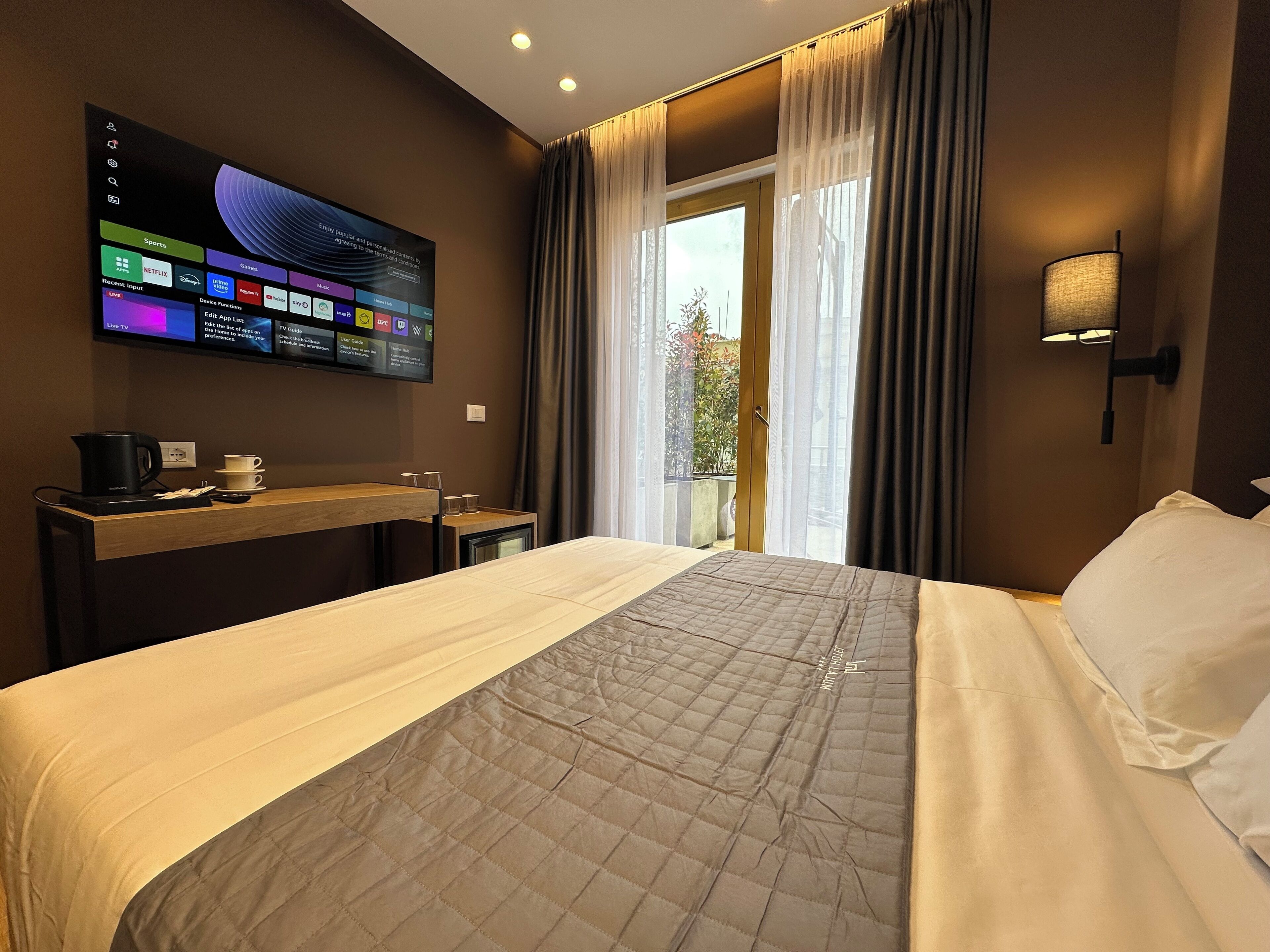 Quarto de hotel com uma cama grande, televisão de tela plana na parede, cafeteira e vista para o exterior por uma porta de correr.