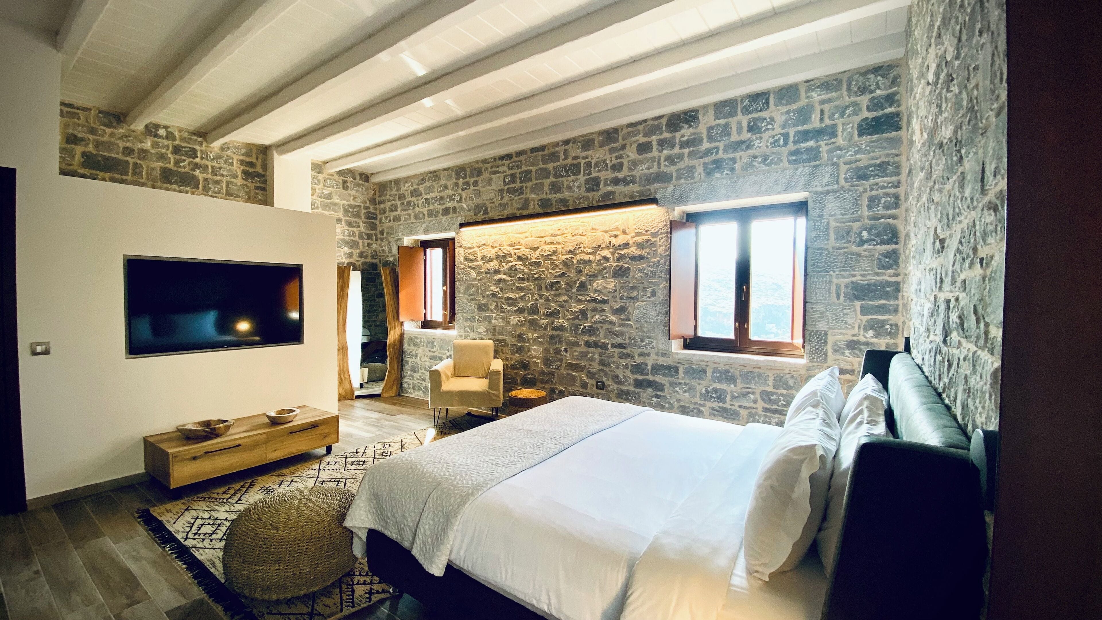 Quarto com parede de pedra, cama, televisão e poltrona.