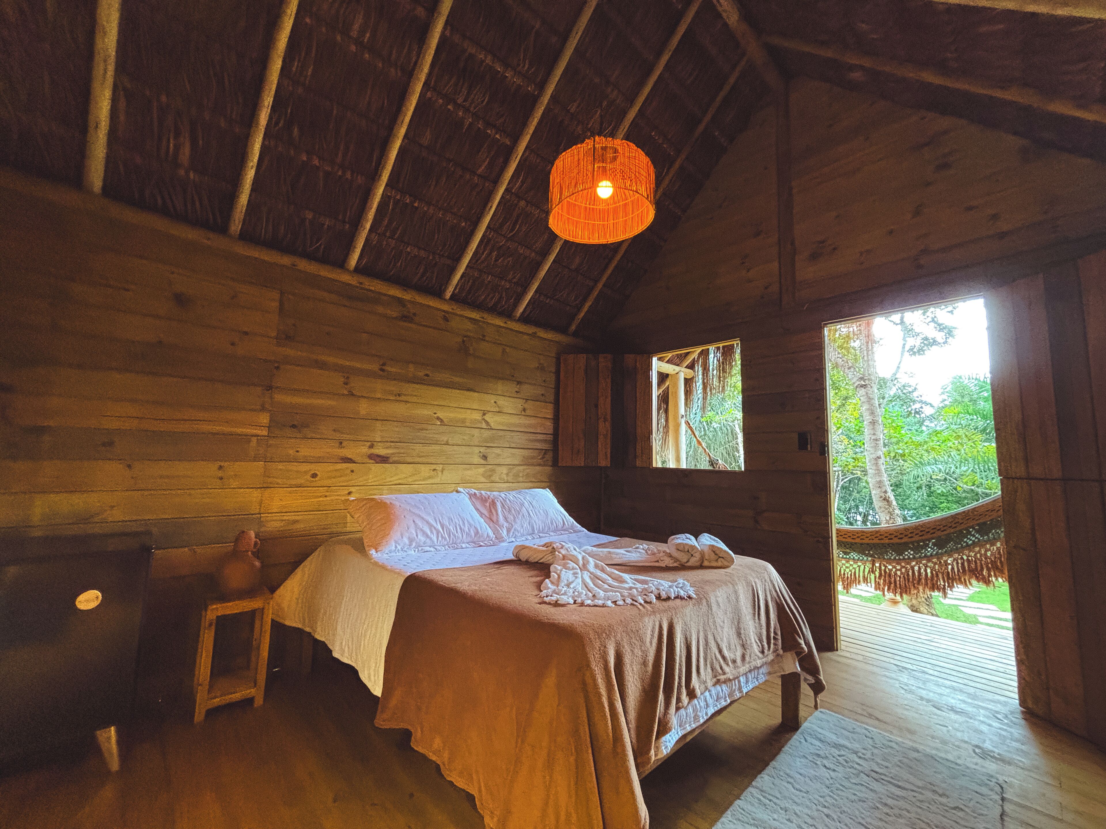 Quarto de cabana de madeira com uma cama, uma mesa pequena, luminária pendente e janela com vista para vegetação.