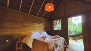 Quarto de cabana de madeira com uma cama, uma mesa pequena, luminária pendente e janela com vista para vegetação.