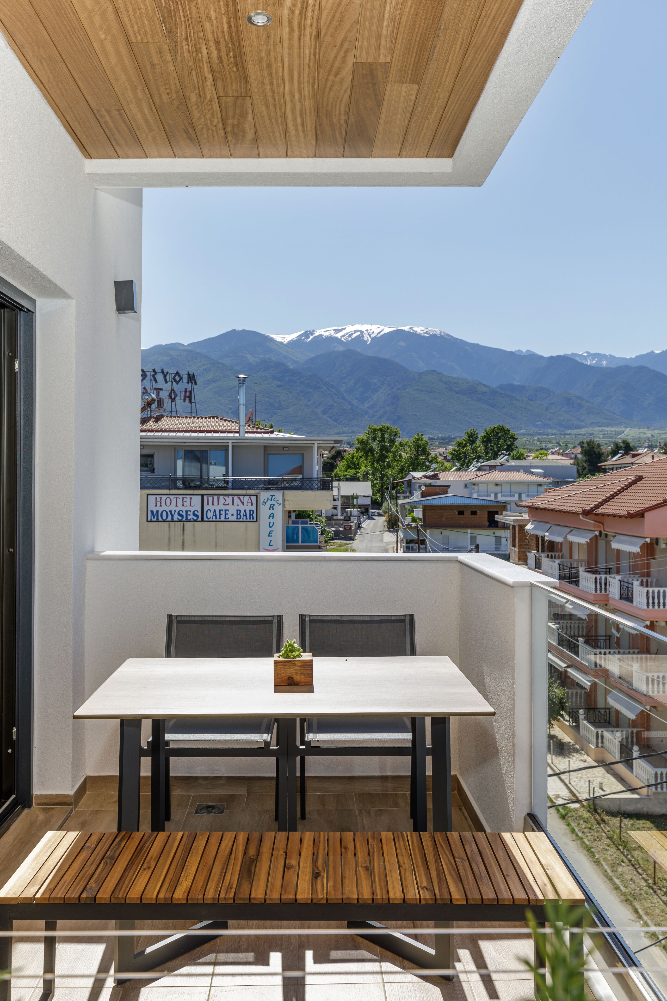 Uma varanda com mesa e banco de madeira, com vista para uma cidade, edifícios e montanhas ao longe.