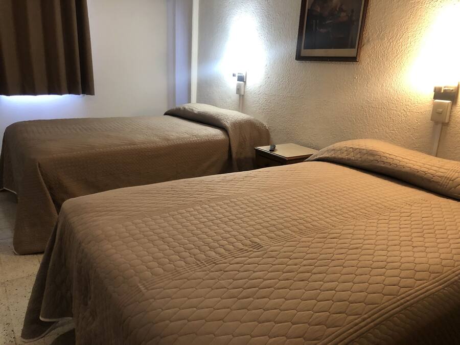 Quarto de hotel com duas camas cobertas por colchas marrons, uma mesa de cabeceira com um controle remoto e uma luminária fixada na parede.