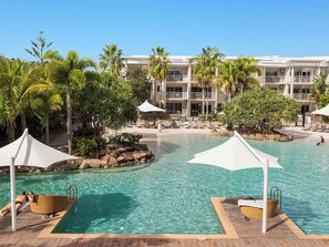 Uma área de piscina em resort com água cristalina, cercada por palmeiras e guarda-sóis, e um edifício com varandas ao fundo.
