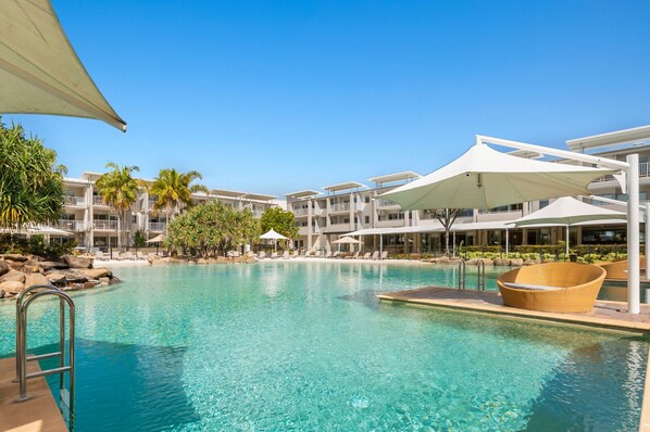 Piscina de resort com águas cristalinas, rodeada por espreguiçadeiras e guarda-sóis, e edifícios modernos ao fundo.