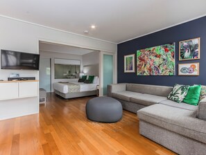 Quarto de hotel moderno com uma cama grande, um sofá cinzento, um banquinho cinzento e uma televisão de tela plana em um armário branco.