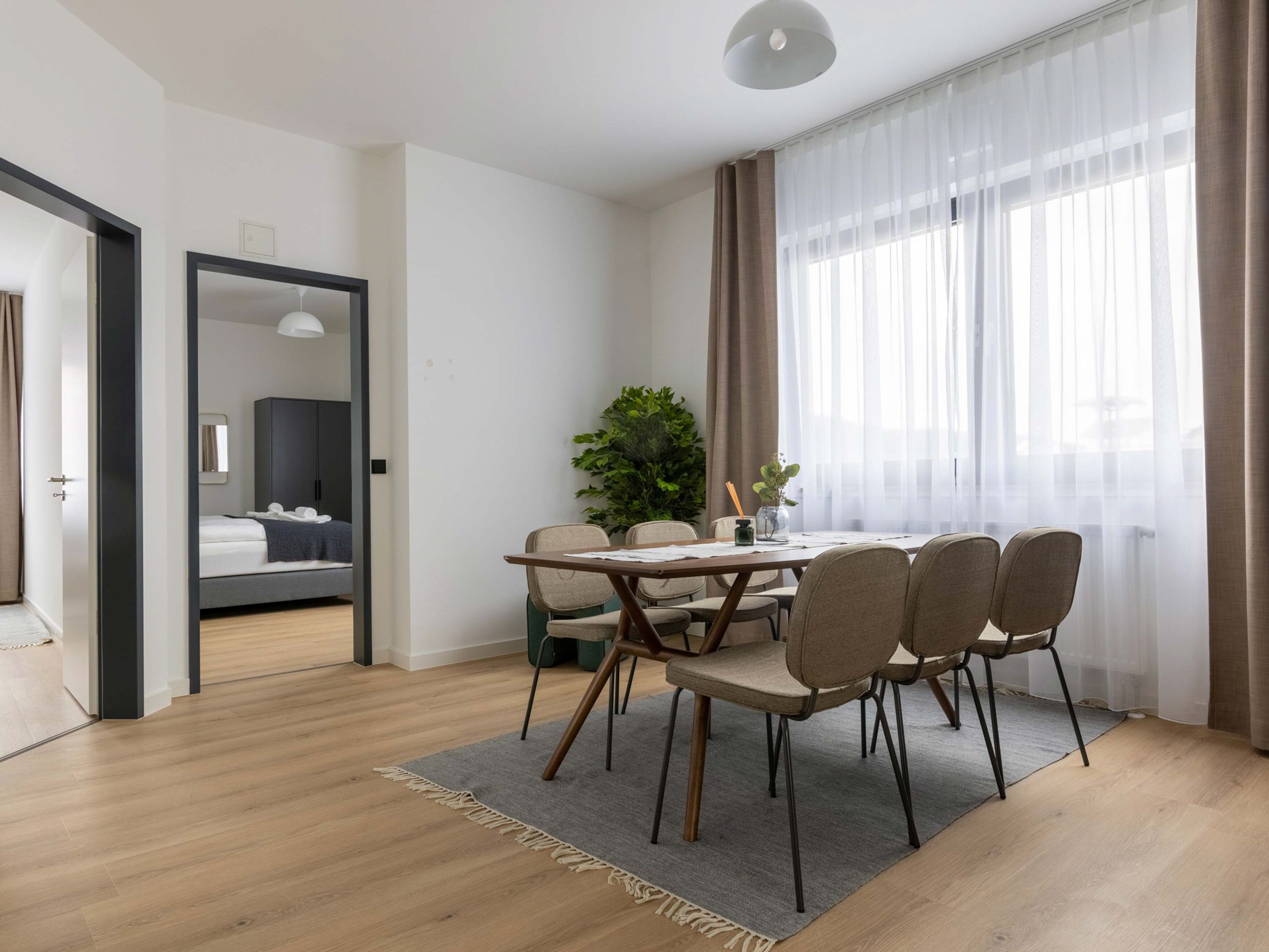 Quarto de hotel moderno com área de jantar, cama, espelho e uma janela grande com cortinas.