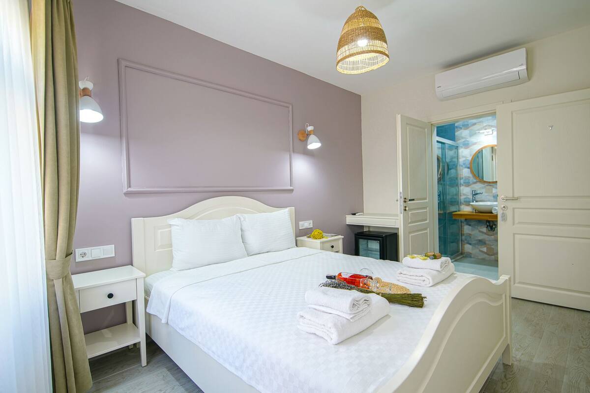 Cama bem arrumada com lençóis brancos, mesa de cabeceira com abajur e um banheiro visível por uma porta aberta.