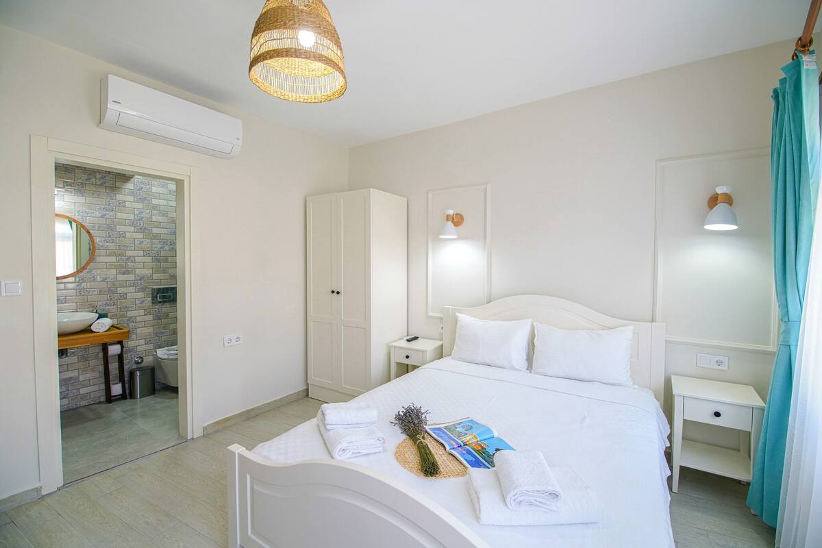 Quarto de hotel com cama, mesa de cabeceira, armário, banheiro com espelho e luminária pendente.