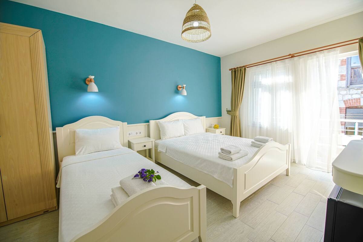 Quarto de hotel com duas camas, um guarda-roupa, uma escrivaninha e janela com cortinas.