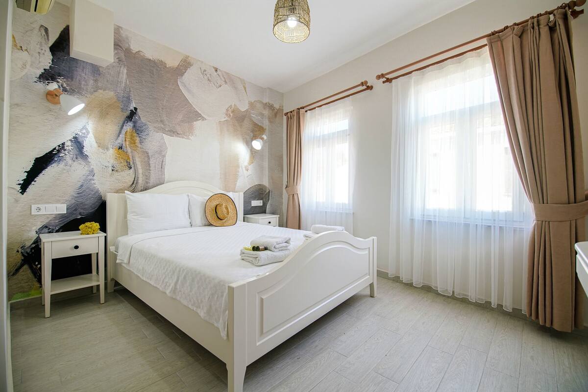 Um quarto com cama, mesas de cabeceira, parede com arte abstrata, janela com cortinas e luminária suspensa.