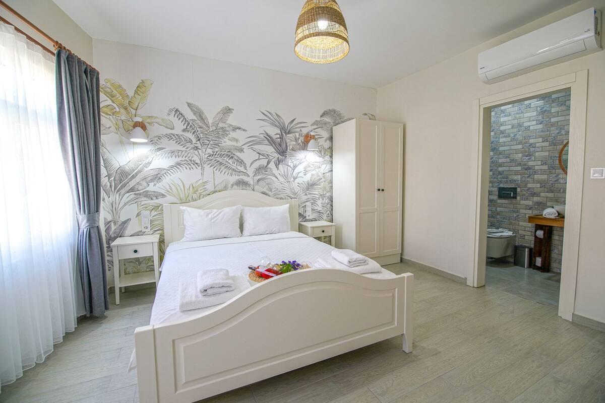 Quarto com cama, mesas de cabeceira, guarda-roupa e banheiro visível por uma porta aberta.