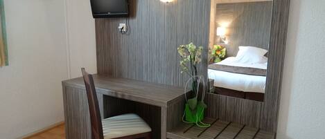 Quarto de hotel compacto com secretária de madeira, uma cadeira, televisão de tela plana presa na parede, mesa de cabeceira com espelho e cama com roupa de cama branca.