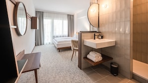 Quarto de hotel com uma cama, uma escrivaninha, uma cadeira, um espelho redondo, uma pia e uma pequena prateleira com toalhas.