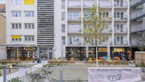 Um edifício moderno de vários andares com varandas, uma pequena árvore em primeiro plano e uma placa anunciando um buffet.