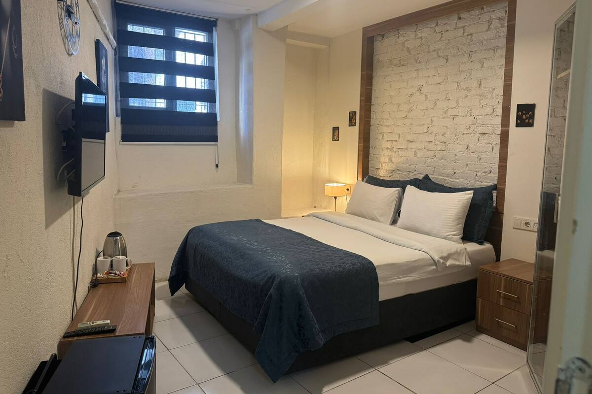 Quarto com cama, mesas de cabeceira, televisão e janela com persianas.