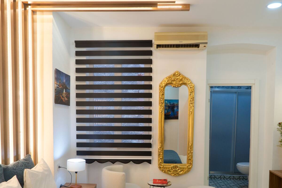Quarto moderno com uma cama grande, mesa de cabeceira com abajur, espelho e janela com persianas.