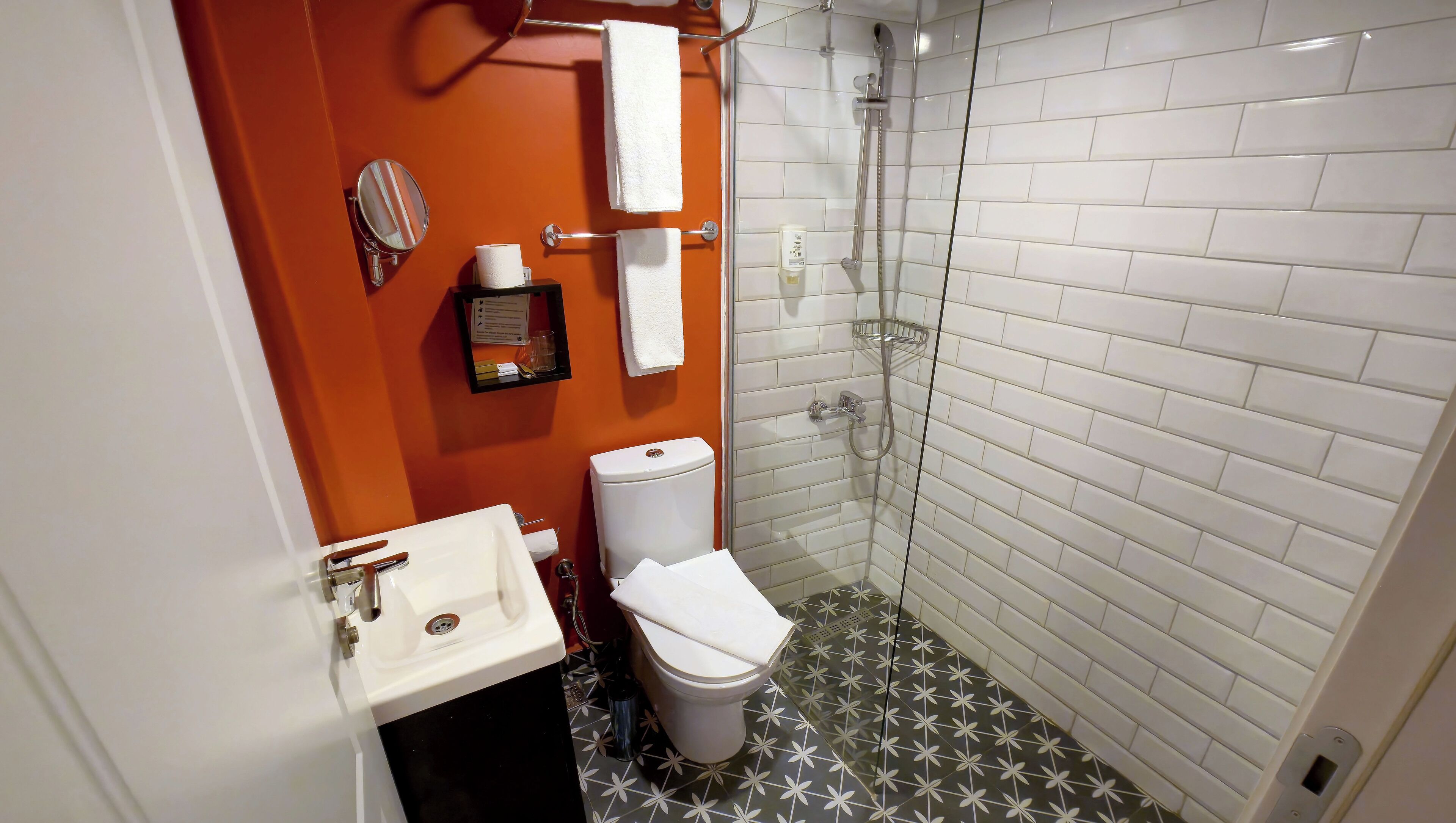 Casa de banho com lavatório branco, sanita e chuveiro. Há um espelho, um porta-toalhas e uma pequena estante com artigos de higiene pessoal. O chão tem um padrão de azulejos.