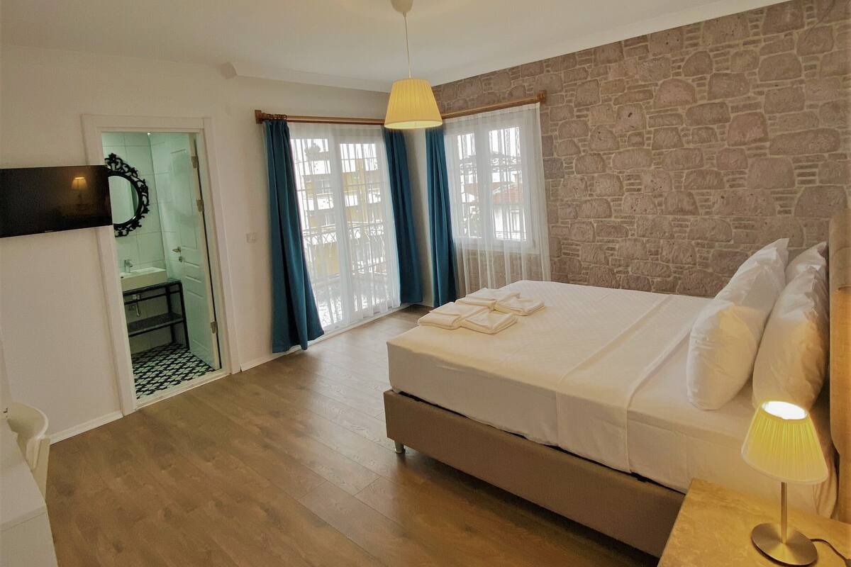 Quarto com cama, mesa de cabeceira com abajur, parede de pedra, janela com cortinas e porta que dá para um banheiro.