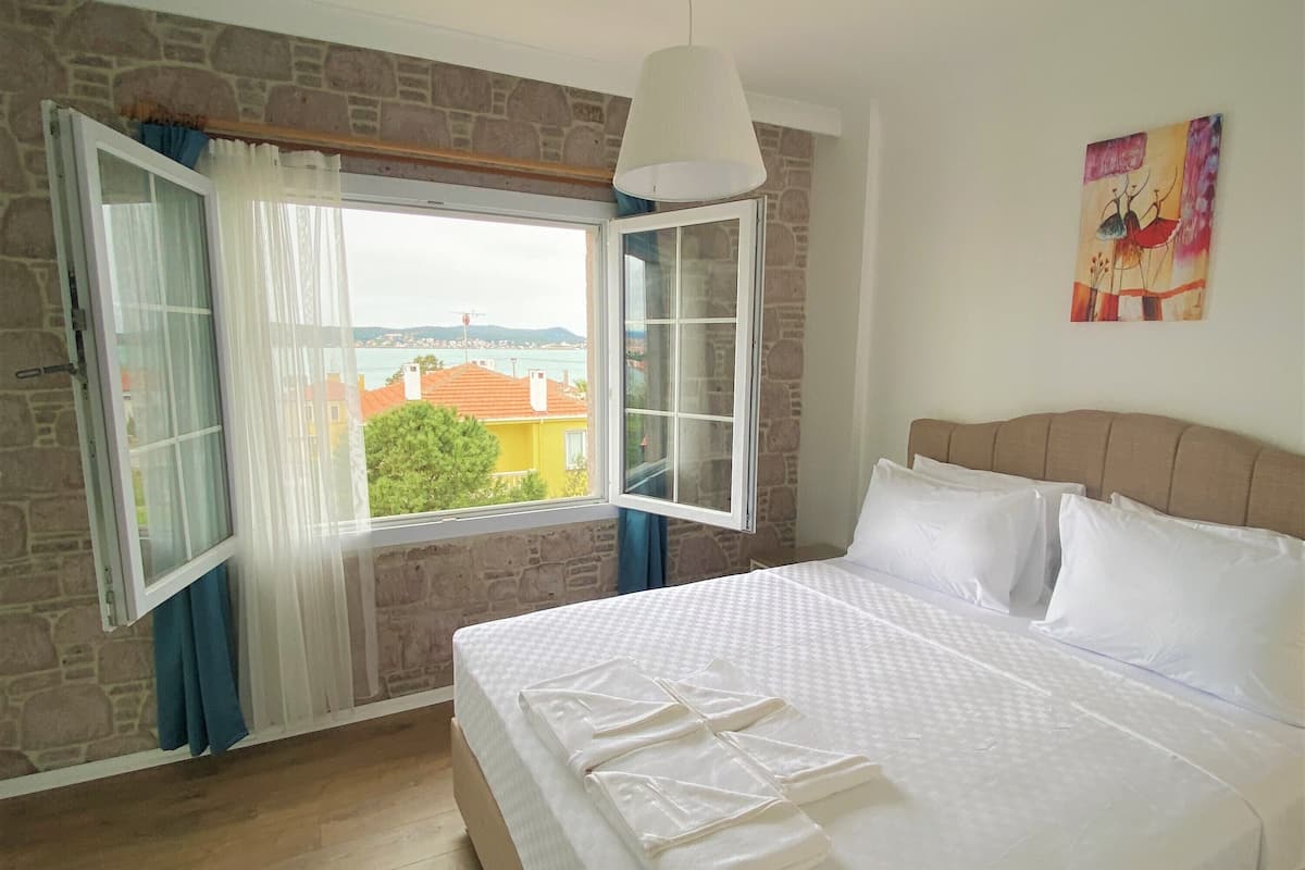 Quarto com cama grande, vista da cidade por uma janela aberta e um quadro na parede.