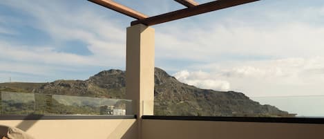 Terraço ensolarado com espreguiçadeiras e uma mesa, com vista para uma paisagem montanhosa.