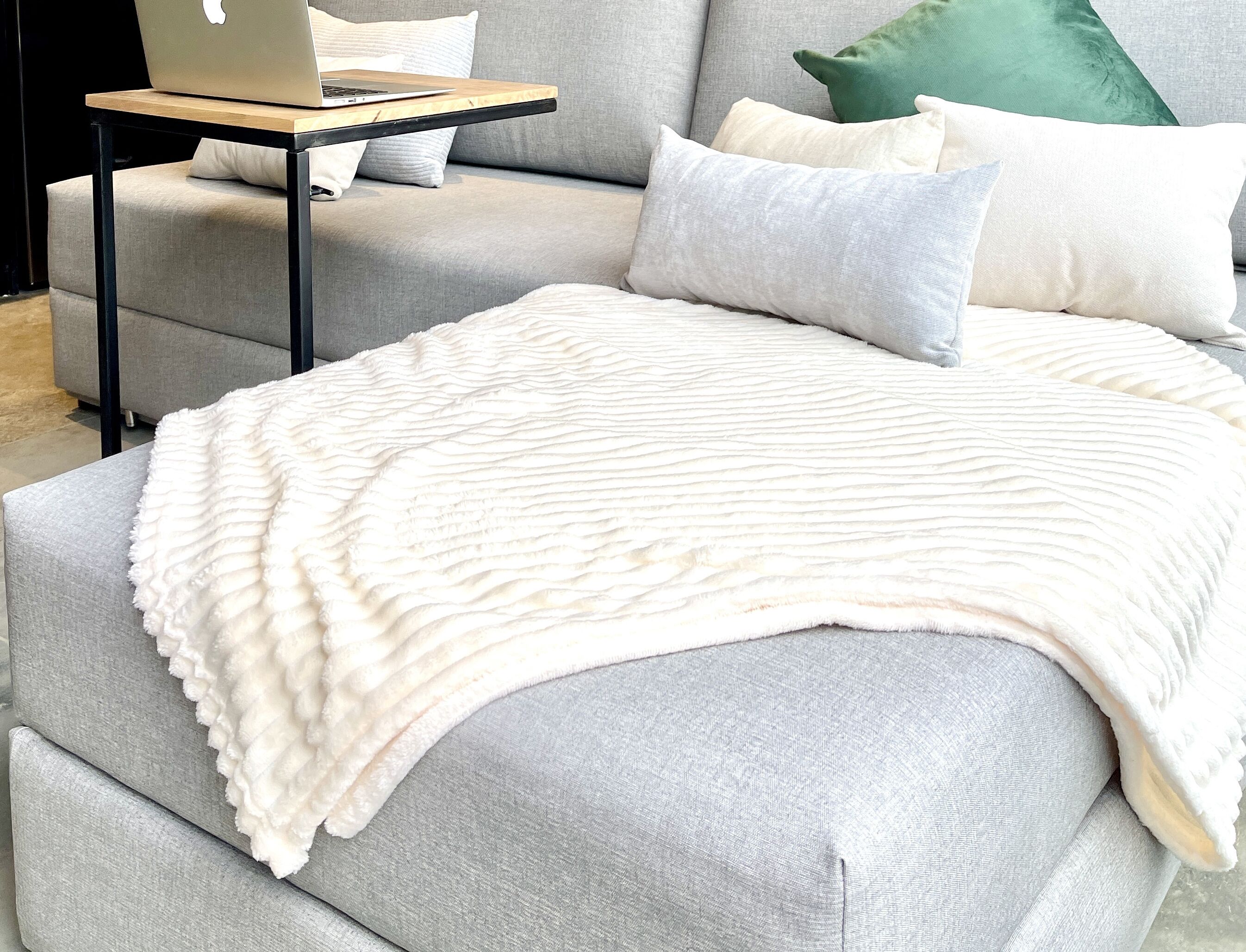 Uma cama com um cobertor branco texturizado, um laptop sobre uma mesinha de madeira e um sofá cinza com almofadas.