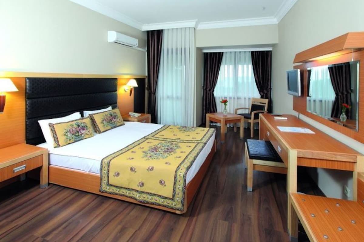 Quarto de hotel com cama grande, escrivaninha de madeira, cadeira, televisão e espelho.