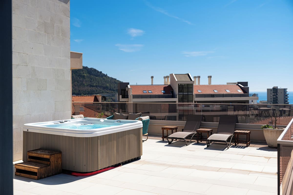 Um terraço no telhado com hidromassagem, espreguiçadeiras e vista para edifícios e colinas.