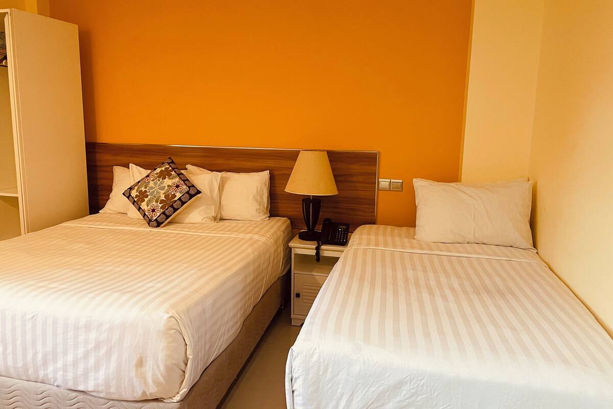 Quarto de hotel com duas camas, uma mesa de cabeceira com luminária e telefone, e parede laranja.