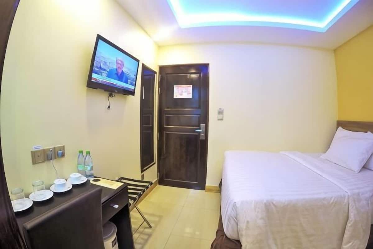 Quarto de hotel com uma cama, uma escrivaninha com cadeira, uma televisão na parede e uma porta.