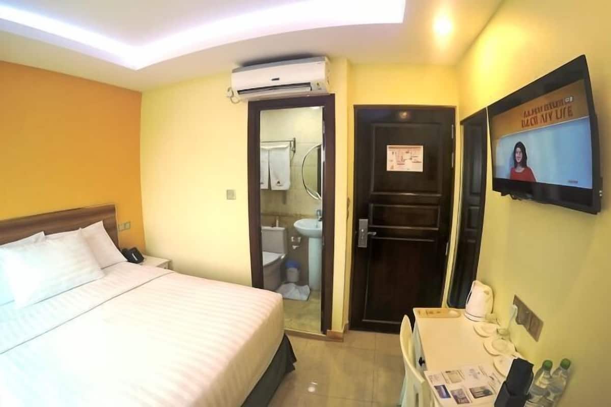Quarto de hotel com uma cama, uma escrivaninha com cadeira, uma televisão e uma porta que dá para um banheiro.