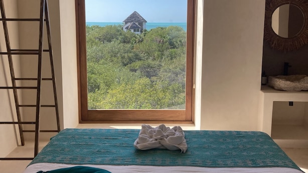 Quarto com uma janela grande, cama com roupa de cama verde-água, escada e vista para uma casa e vegetação.