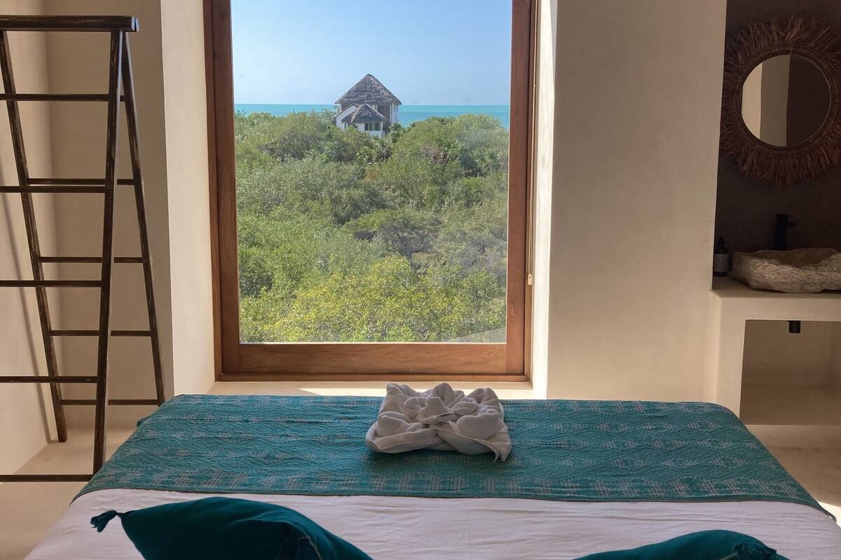 Quarto com uma janela grande, cama com roupa de cama verde-água, escada e vista para uma casa e vegetação.