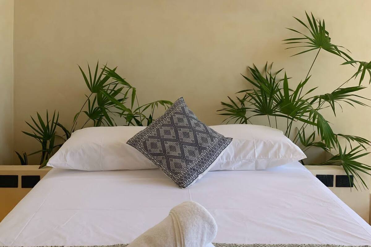 Uma cama bem arrumada com um travesseiro estampado, uma janela acima e plantas verdes ao redor.