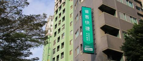 Edifício de vários andares com fachada verde e cinza, apresentando varandas e janelas. Uma placa verde com texto branco indica um hotel chamado "Fengchuan" e menciona "CHECKIN Express".