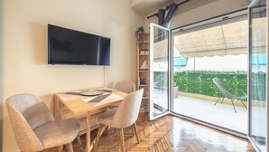 Quarto moderno com piso em padrão herringbone de madeira, TV de tela plana na parede, mesa de jantar com duas cadeiras estampadas e porta de vidro deslizante que dá para uma varanda com vista para um conjunto de arbustos verdes e céu azul.