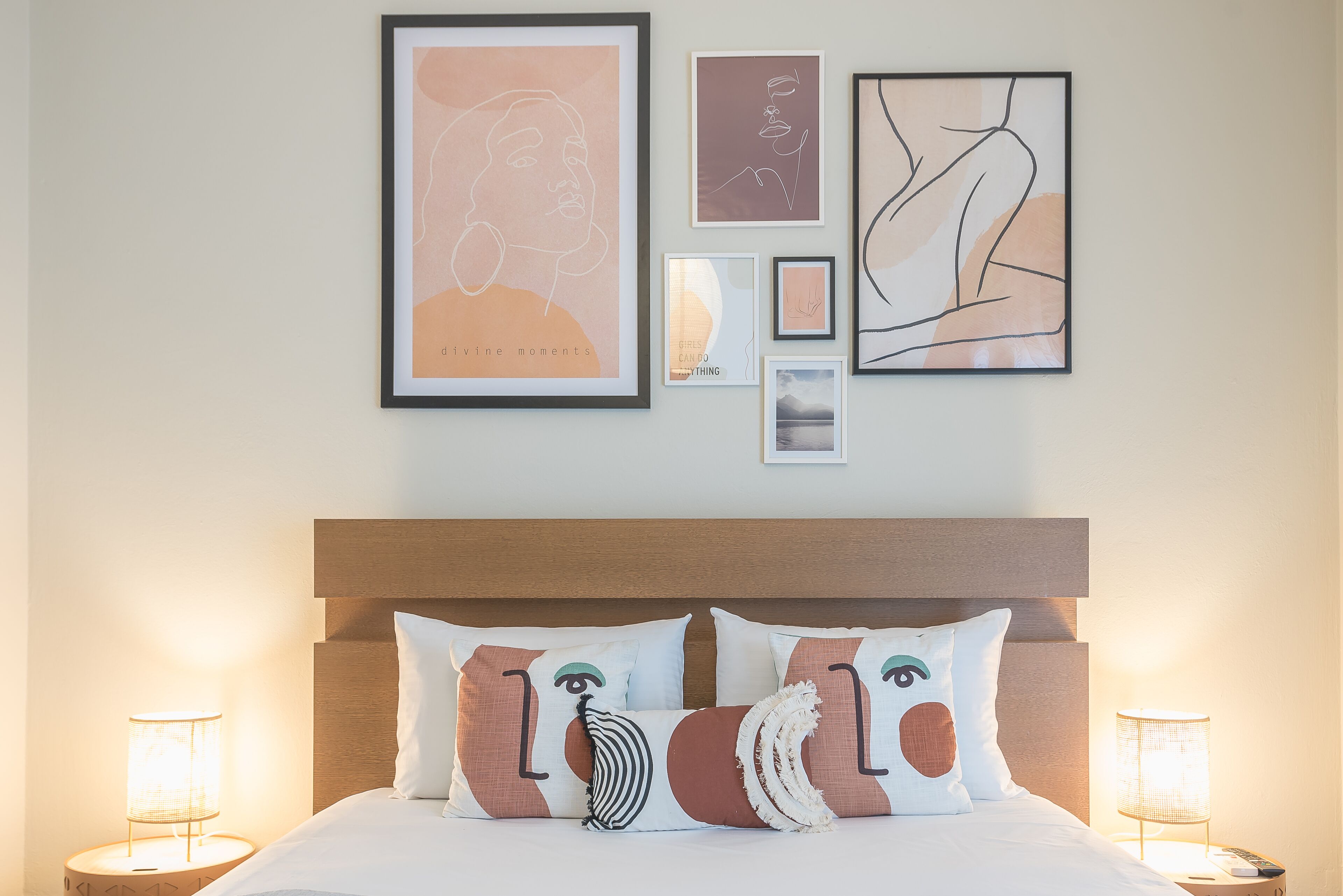 Cama bem arrumada com cabeceira de madeira, duas luminárias de apoio e parede decorada com obras emolduradas.