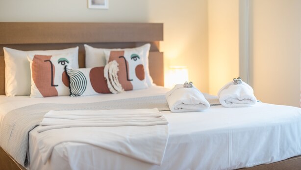 Cama bem arrumada com lençóis brancos e almofadas decorativas, uma luminária de cabeceira e uma toalha dobrada na mesinha de apoio.