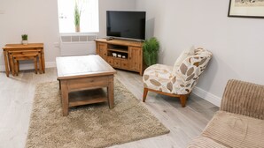 Uma sala de estar com uma mesa de centro de madeira, um sofá, um rack para TV e uma pequena mesa com uma planta.