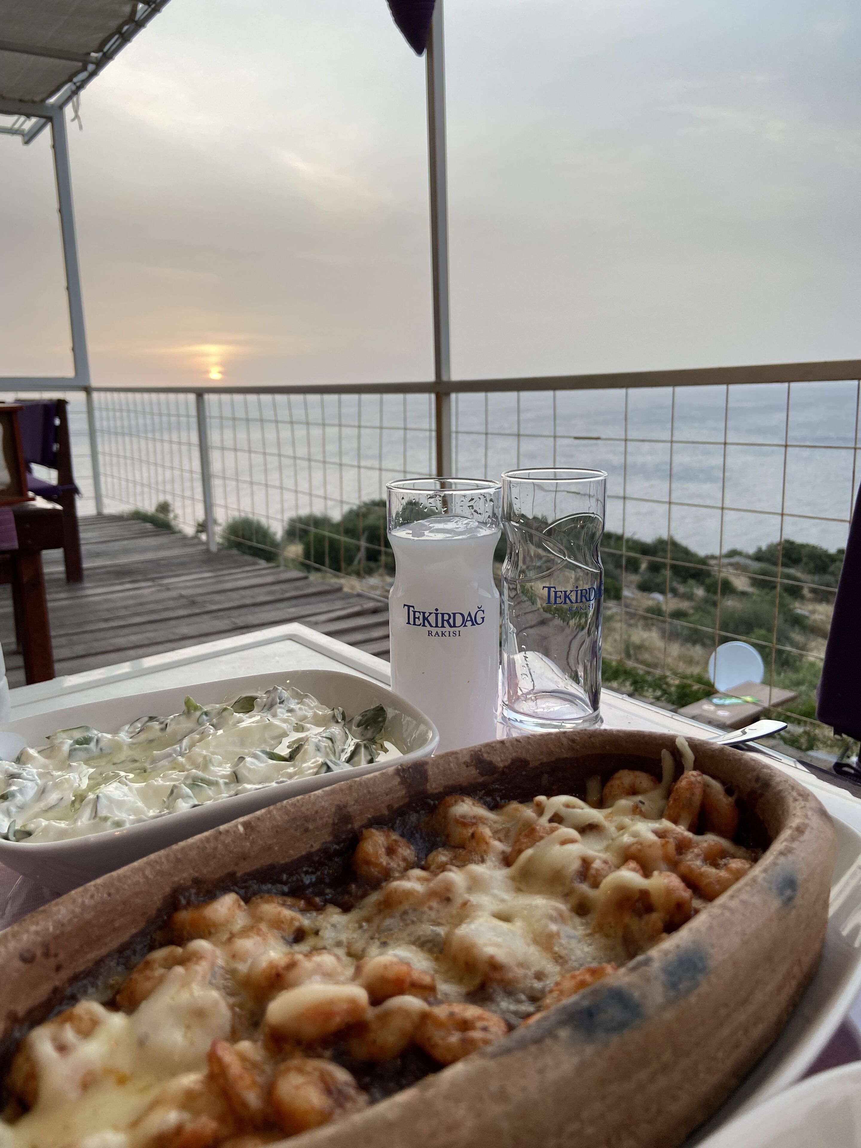 Uma mesa de jantar com um prato de massa, uma saladeira, uma garrafa de Tekirdağ Rakisi e dois copos de água, dispostos numa varanda com vista para o mar e dunas.
