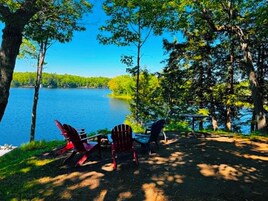 Um camping à beira do lago com duas cadeiras Adirondack e uma mesa, cercado por árvores e uma vista desobstruída do lago.