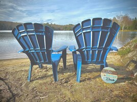 Duas cadeiras Adirondack azuis de frente para um lago, com uma placa que diz 'A vida é melhor na praia'.