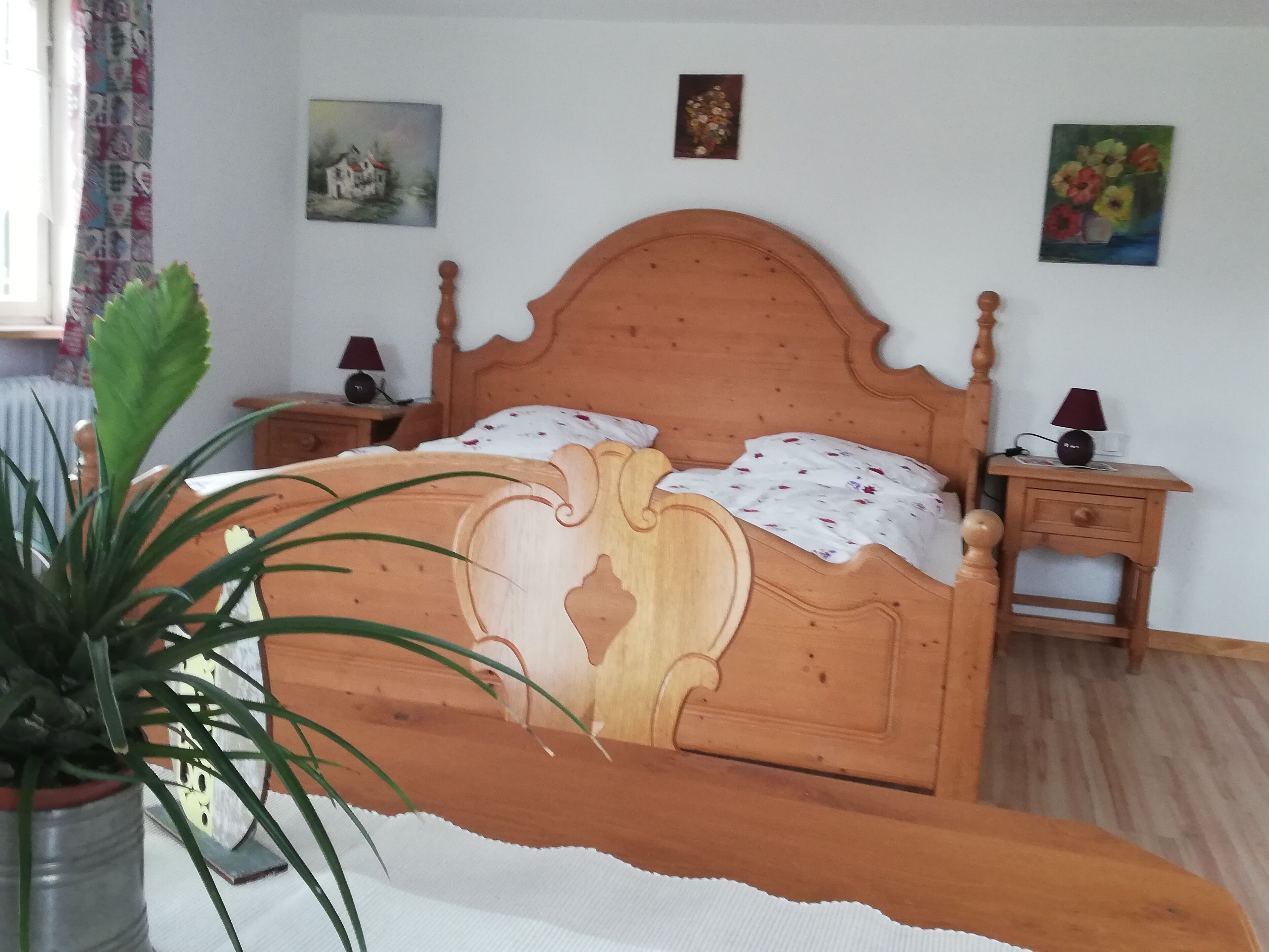 Quarto com cama de madeira, duas mesas de cabeceira, uma planta em vaso e dois quadros pendurados na parede.