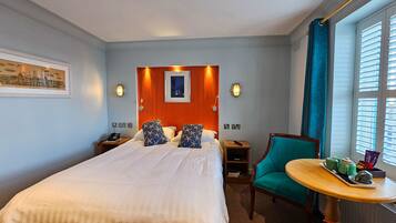 Quarto de hotel com uma cama grande, cabeceira de madeira, duas mesas de cabeceira, uma poltrona verde, uma mesinha redonda com bandeja e uma janela com persianas.