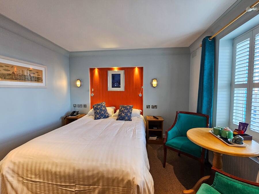 Quarto de hotel com uma cama grande, cabeceira de madeira, duas mesas de cabeceira, uma poltrona verde, uma mesinha redonda com bandeja e uma janela com persianas.