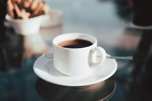 Uma chávena de café branco num pires, com uma colher, pousada sobre uma superfície refletora.