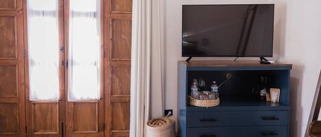 Quarto com porta de madeira, televisão sobre um armário azul e um cesto com garrafas e copos.
