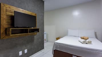 Quarto de hotel com uma cama, uma televisão presa a um painel de madeira e uma pequena mesa de cabeceira.