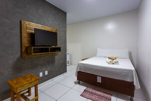 Quarto de hotel com uma cama, televisão fixada na parede, um banquinho de madeira e um tapete no chão.