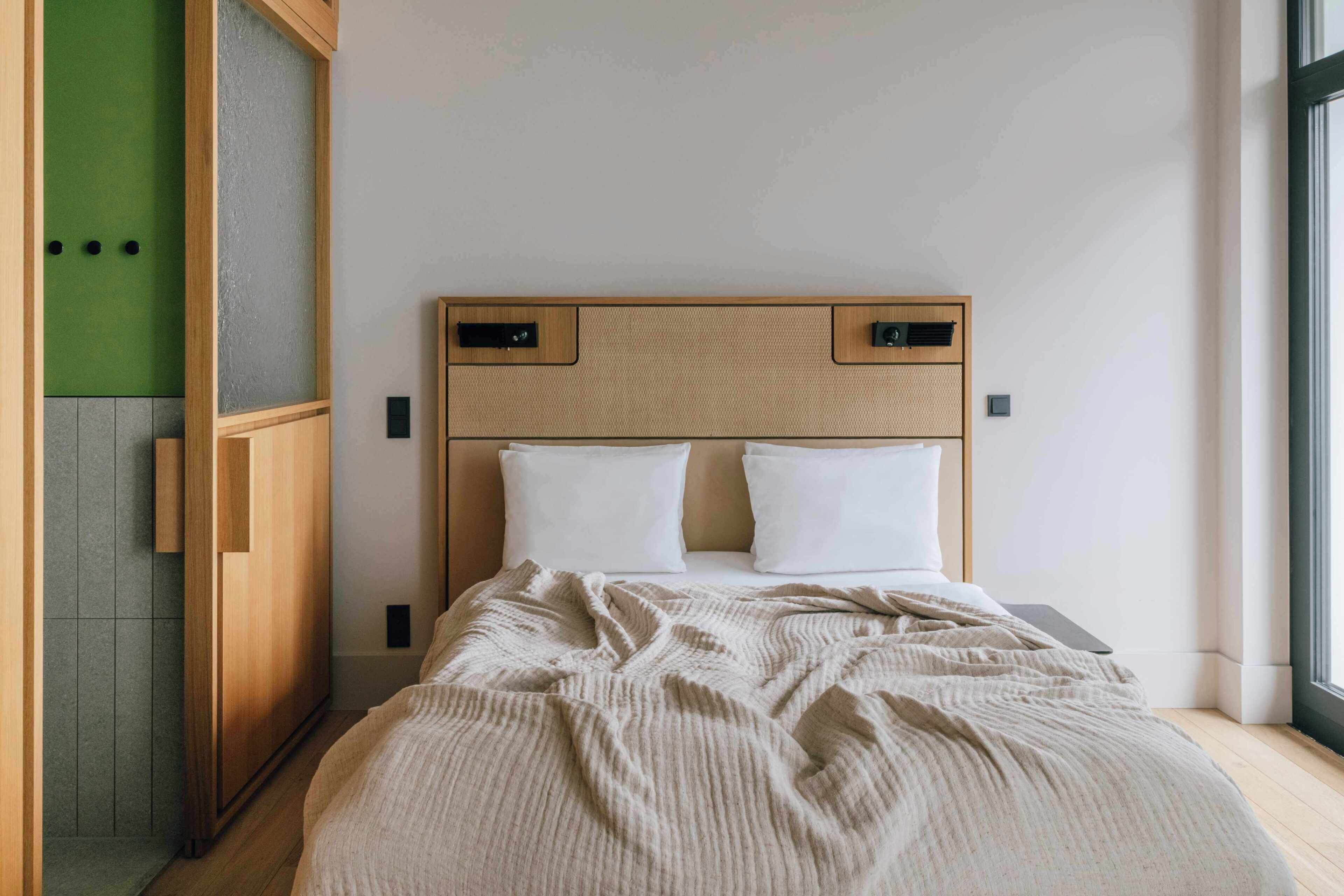 Quarto com cabeceira de madeira, duas almofadas brancas, manta bege e parede verde.